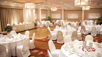 Salão de banquetes com mesas redondas dispostas para um evento formal, com toalhas brancas e cadeiras, e luminárias decorativas.