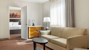 Quarto de hotel com cama, sofá, mesa pequena, luminária e vista para a cidade através de uma janela.