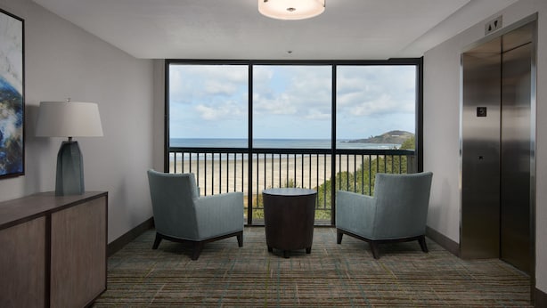 Quarto com duas poltronas, uma mesinha, uma lâmpada e vista para o mar através de janelas amplas.