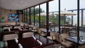 Restaurante com várias mesas e cadeiras, grandes janelas com vista para a praia e uma televisão de tela plana fixada na parede.