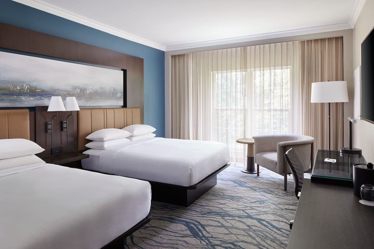 Quarto de hotel com duas camas, uma secretária, uma cadeira, uma janela grande com cortinas, um quadro na parede e um abajur.