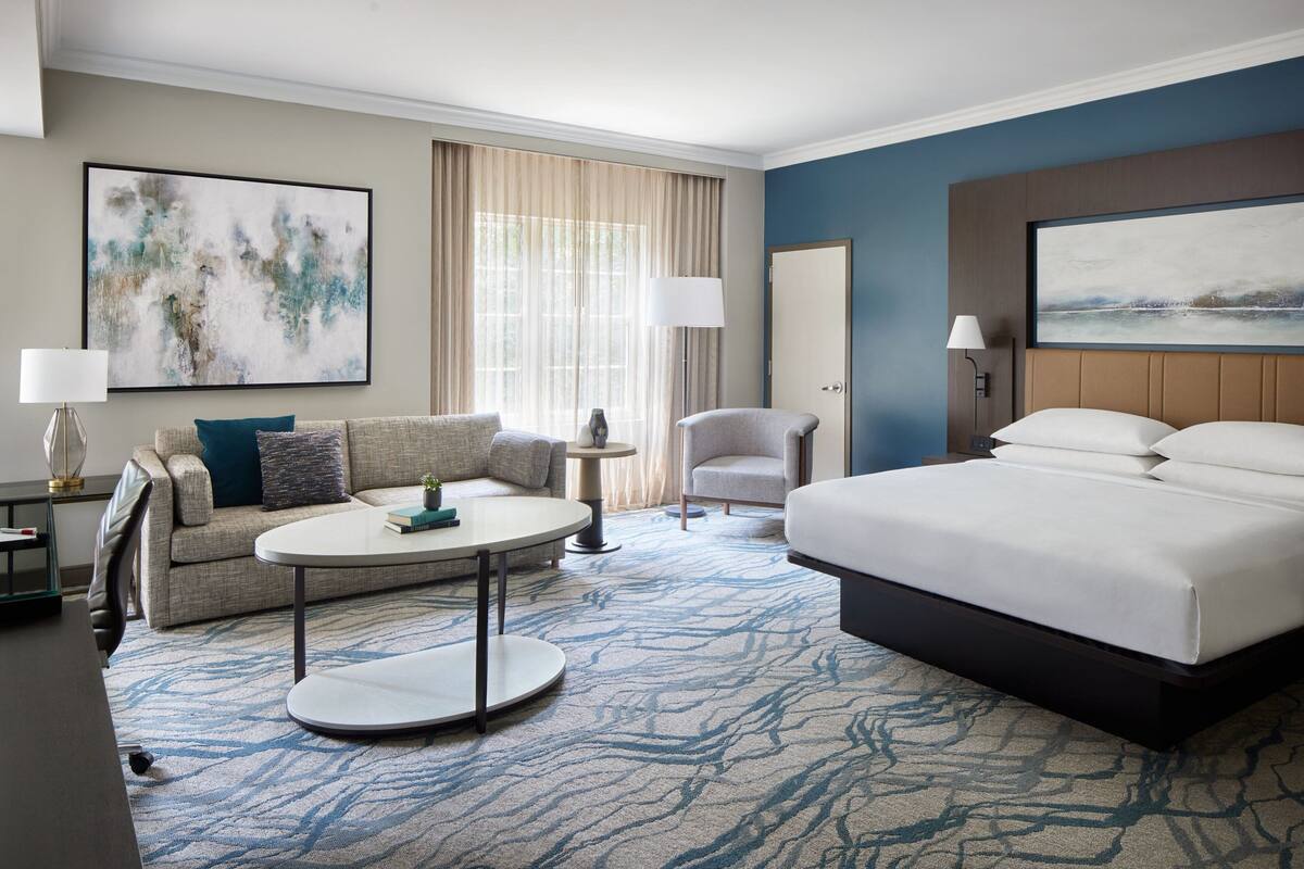 Quarto de hotel com uma cama, um sofá, uma poltrona, uma mesa de centro e um quadro na parede.