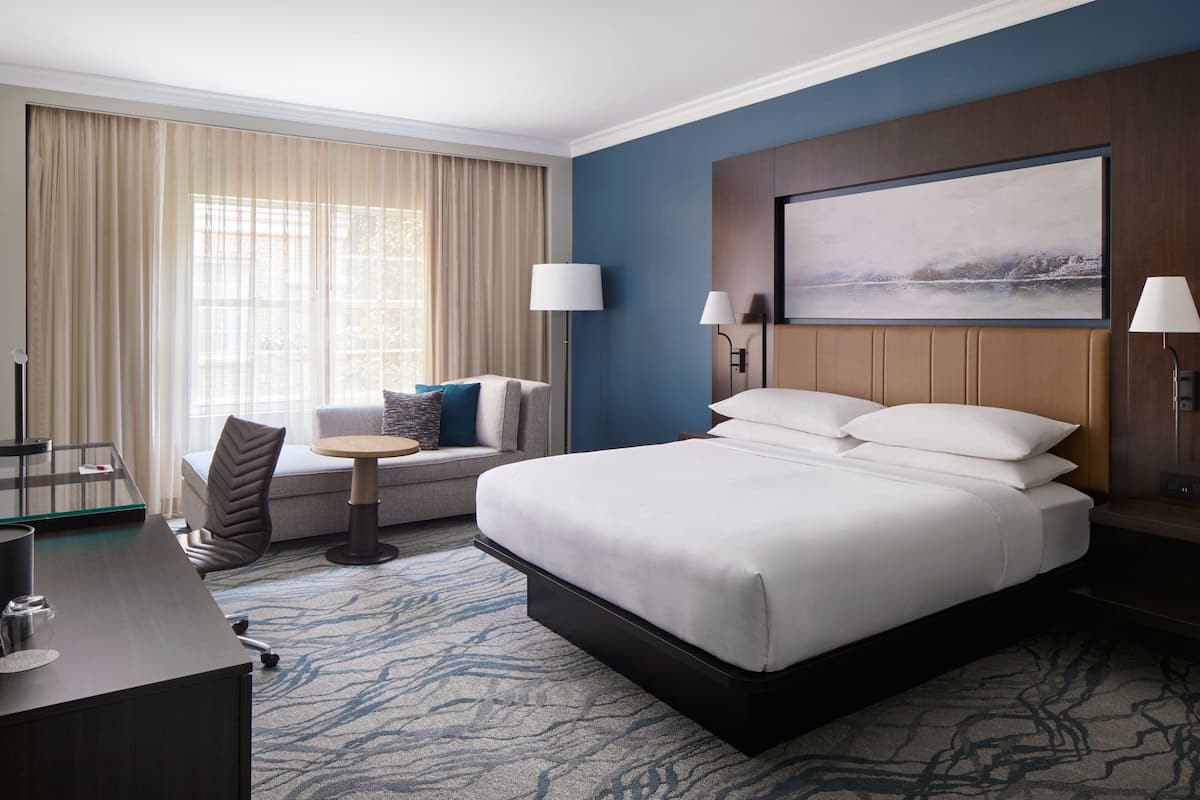 Quarto de hotel com uma cama grande, uma escrivaninha, uma cadeira, um sofá e uma janela ampla com cortinas.