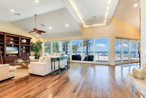 Uma sala de estar ampla com uma grande janela com vista para a praia, piso de madeira e ventoinha no teto.