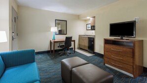 Quarto de hotel com sofá azul, escrivaninha com luminária, televisão, cozinha compacta e minibar.