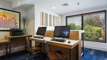 Espaço de escritório moderno com duas mesas, cada uma com monitor de computador, teclado e rato. Há uma impressora à esquerda, e o espaço tem uma janela grande com persianas.