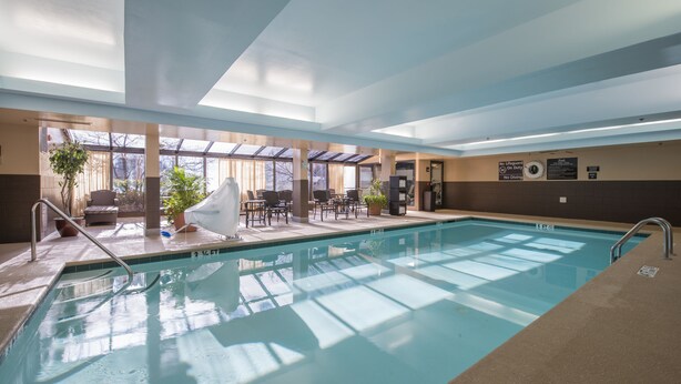 Piscina interior com paredes de vidro transparente, área de estar ao ar livre ensolarada com mesas e cadeiras, e aviso de ausência de salva-vidas.