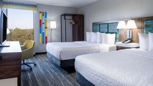 Quarto de hotel com duas camas, uma escrivaninha com computador, uma cadeira e vista para árvores.