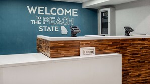 Balcão de recepção com parede azul e parede de destaque em madeira, exibindo uma placa com a mensagem 'Welcome to the Peach State' e uma placa 'Hilton Honors'.