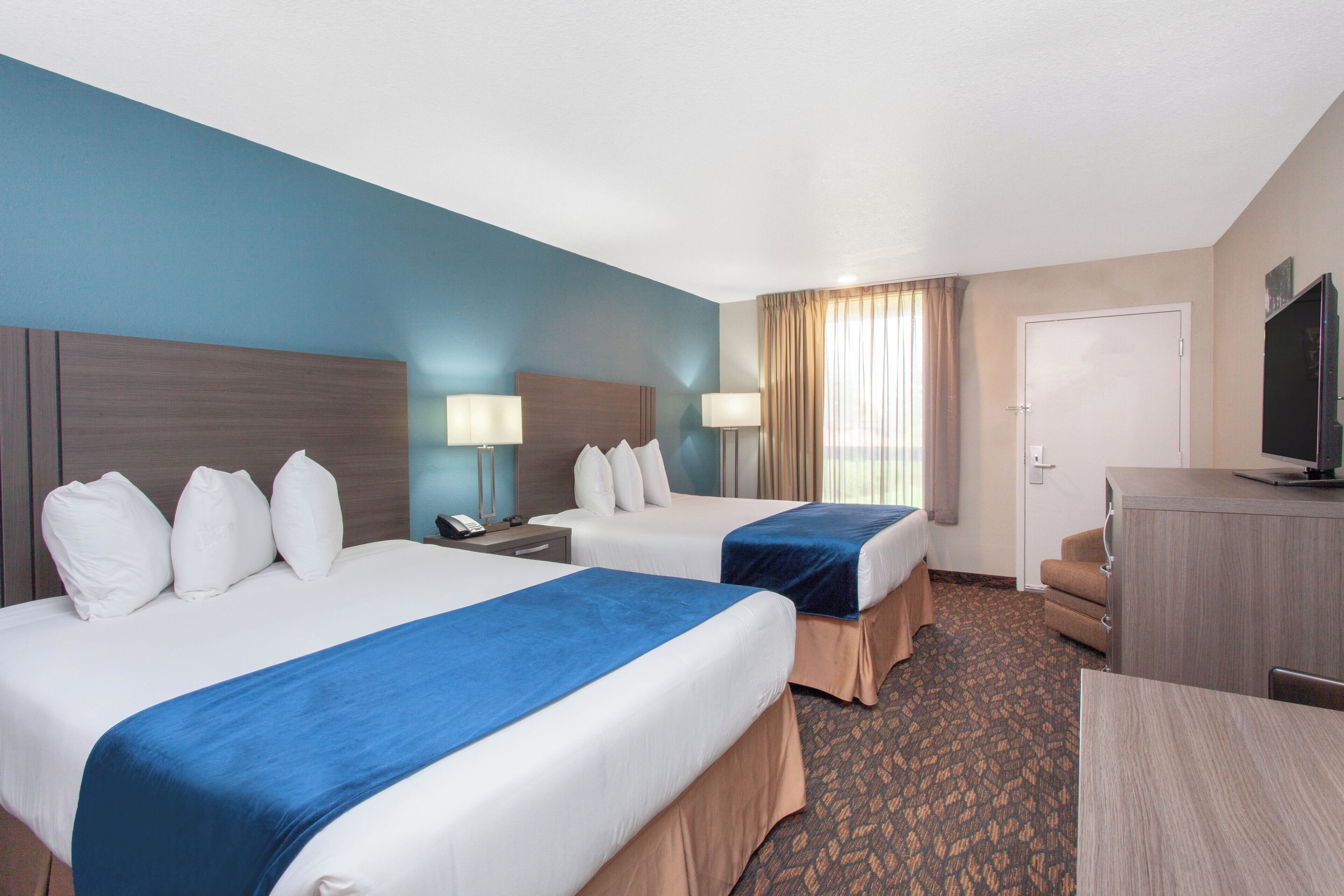 Quarto de hotel com duas camas, uma escrivaninha, uma televisão e uma porta.