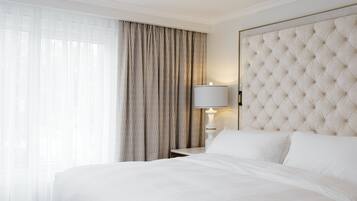 Quarto de hotel com cama grande, mesa de cabeceira com abajur e janela com cortinas leves e cortinas estampadas.