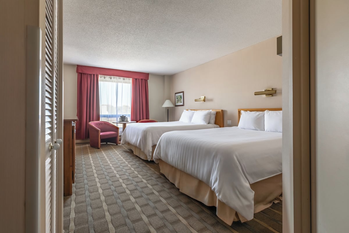 Quarto de hotel com duas camas, uma escrivaninha, uma cadeira e uma janela com cortinas.