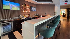Uma área de bar com um balcão comprido, banquetas, uma televisão na parede e uma prateleira com vários frascos.
