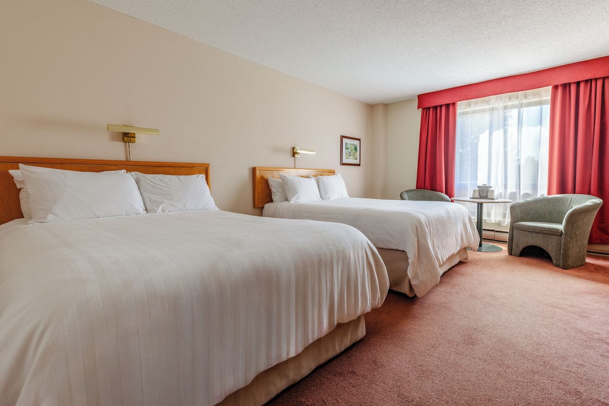 Quarto de hotel com duas camas, uma mesa pequena, uma cadeira e uma janela com cortinas vermelhas.