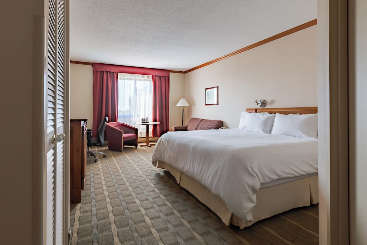 Quarto de hotel com uma cama grande, uma escrivaninha, uma cadeira, um abajur e uma janela com cortinas.