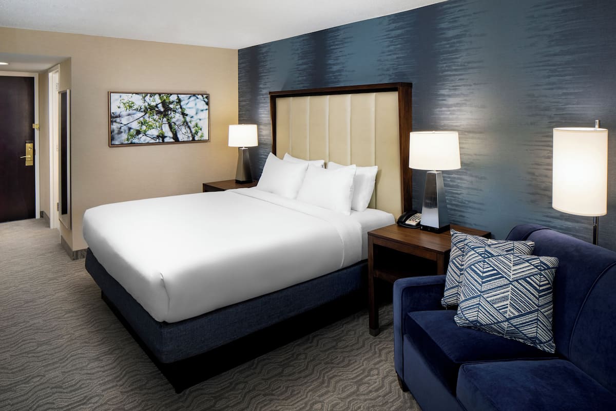 Quarto de hotel com uma cama grande, duas mesas de cabeceira com abajures, um sofá azul escuro com almofadas estampadas e um quadro na parede.