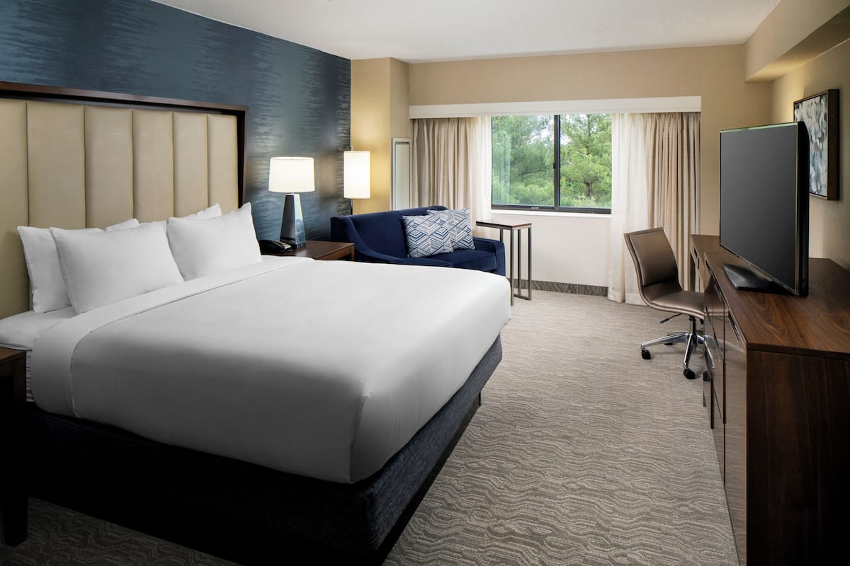 Quarto de hotel com uma cama grande, uma mesa de trabalho com televisão, uma cadeira e uma janela com cortinas.