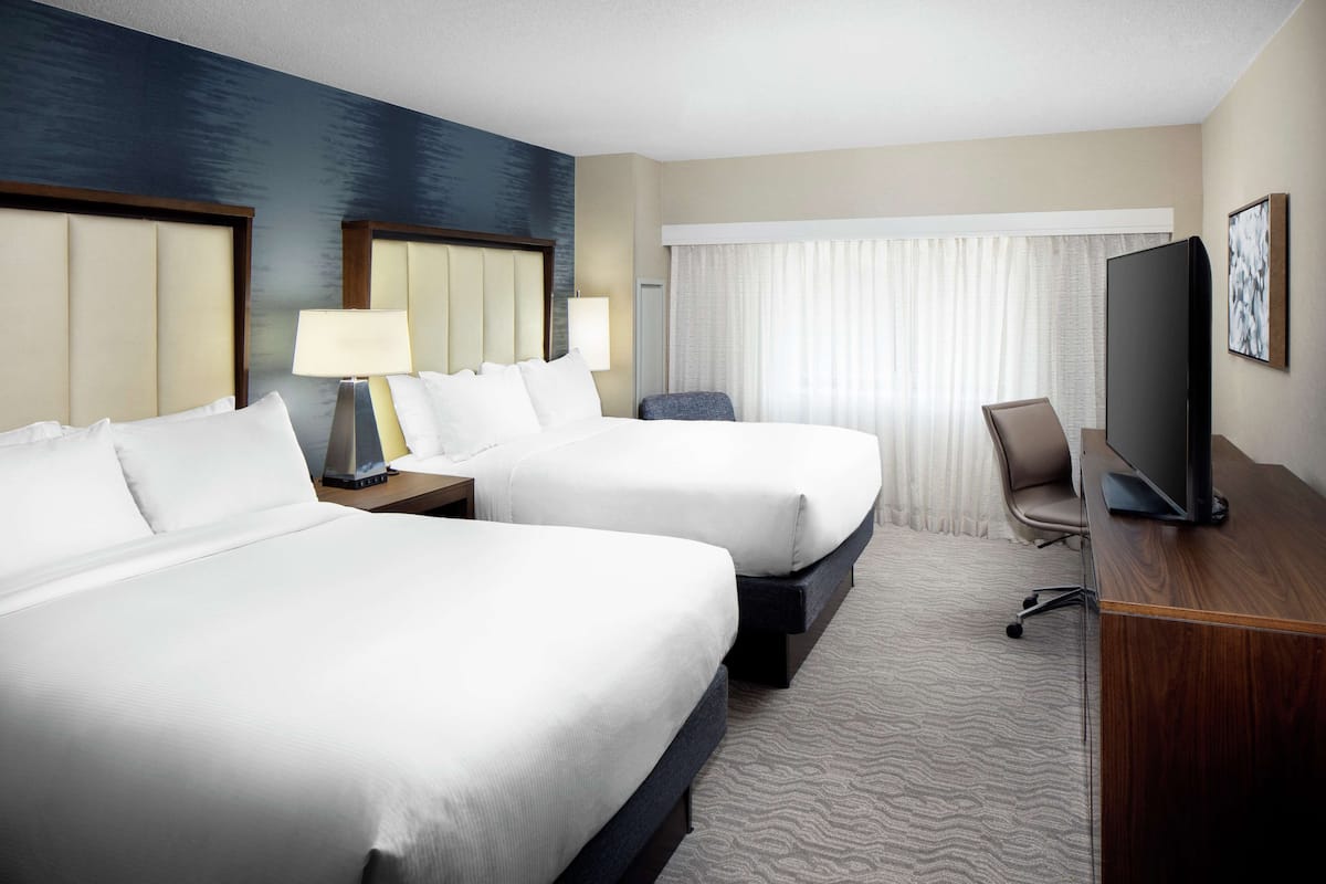 Quarto de hotel com duas camas, uma mesa de trabalho e televisão, e uma janela com cortinas.