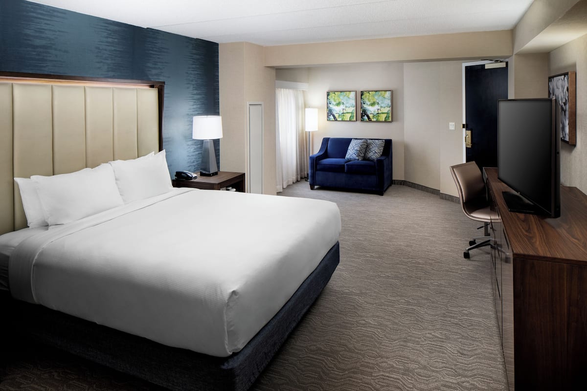 Quarto de hotel com uma cama grande, uma mesa de trabalho com televisão, um sofá e duas pinturas na parede.