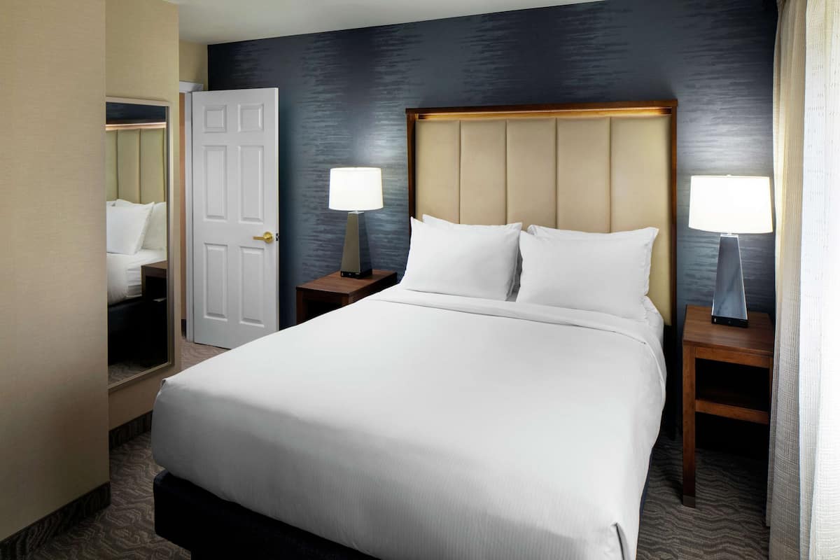 Quarto de hotel com uma cama grande, duas mesas de cabeceira com luminárias, uma porta e um espelho.