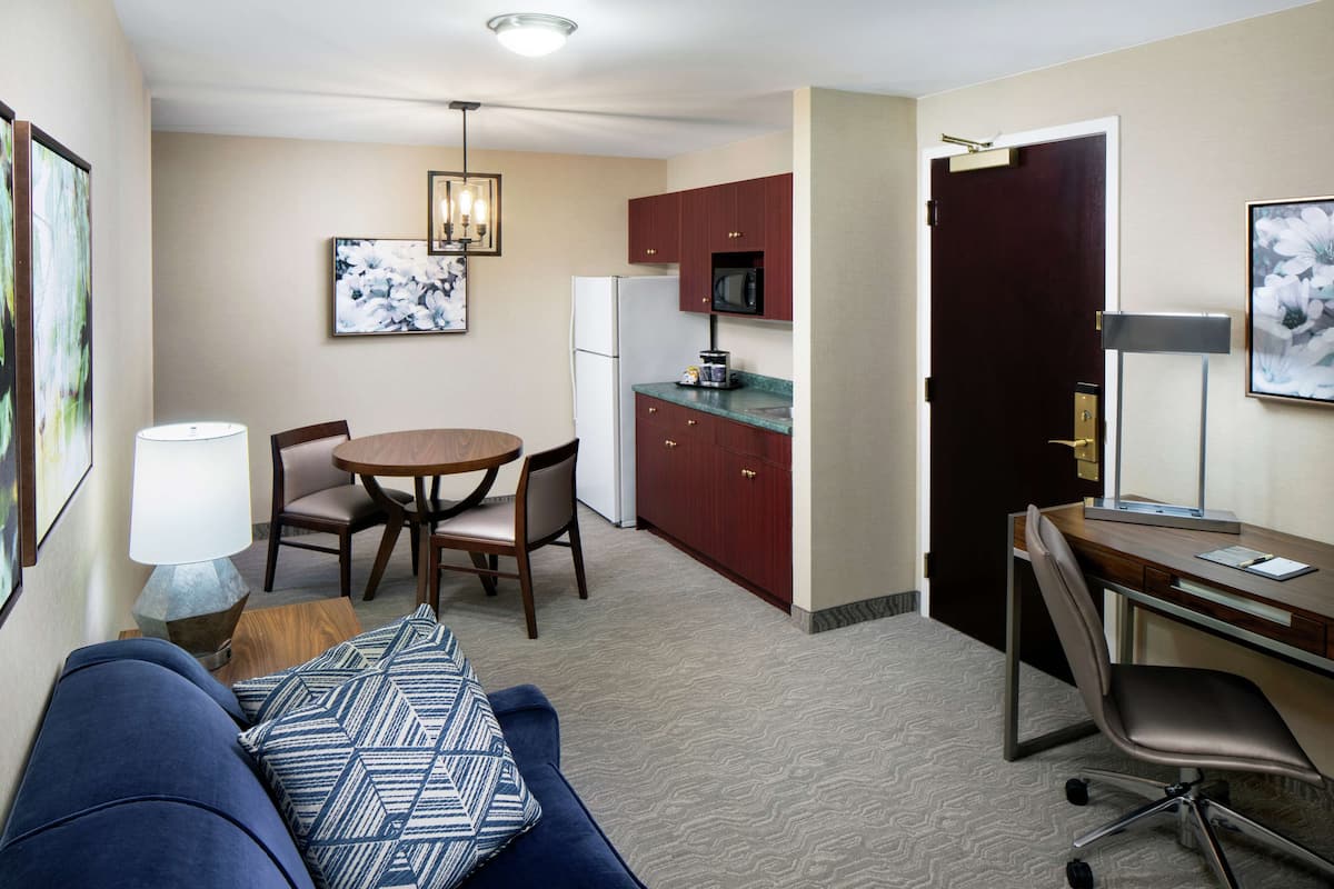 Quarto de hotel compacto com cozinha compacta, área de refeições, sofá com almofadas estampadas e escrivaninha com cadeira.
