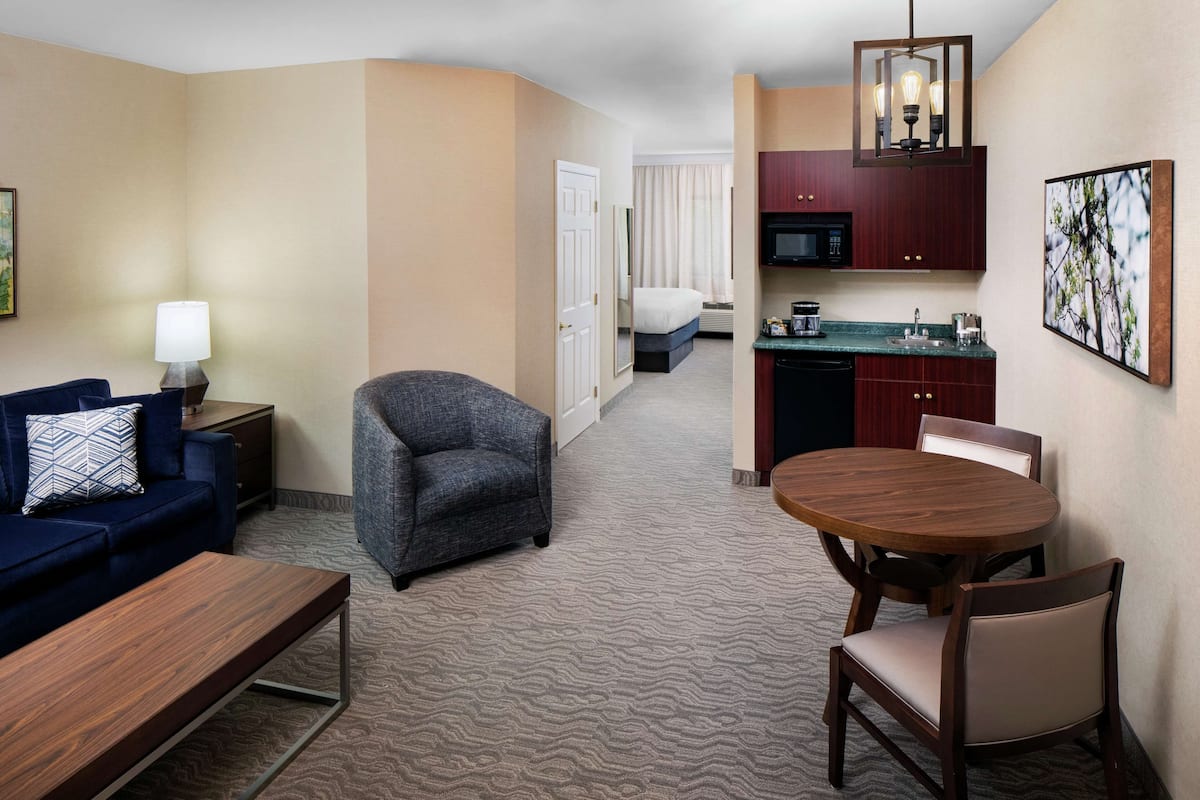 Quarto de hotel com sofá, poltrona, mesa de jantar e cozinha compacta.