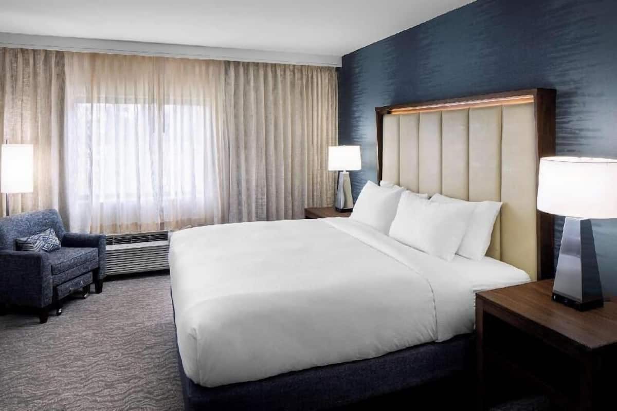 Quarto de hotel com uma cama grande, uma cadeira, uma mesa de cabeceira e um abajur.
