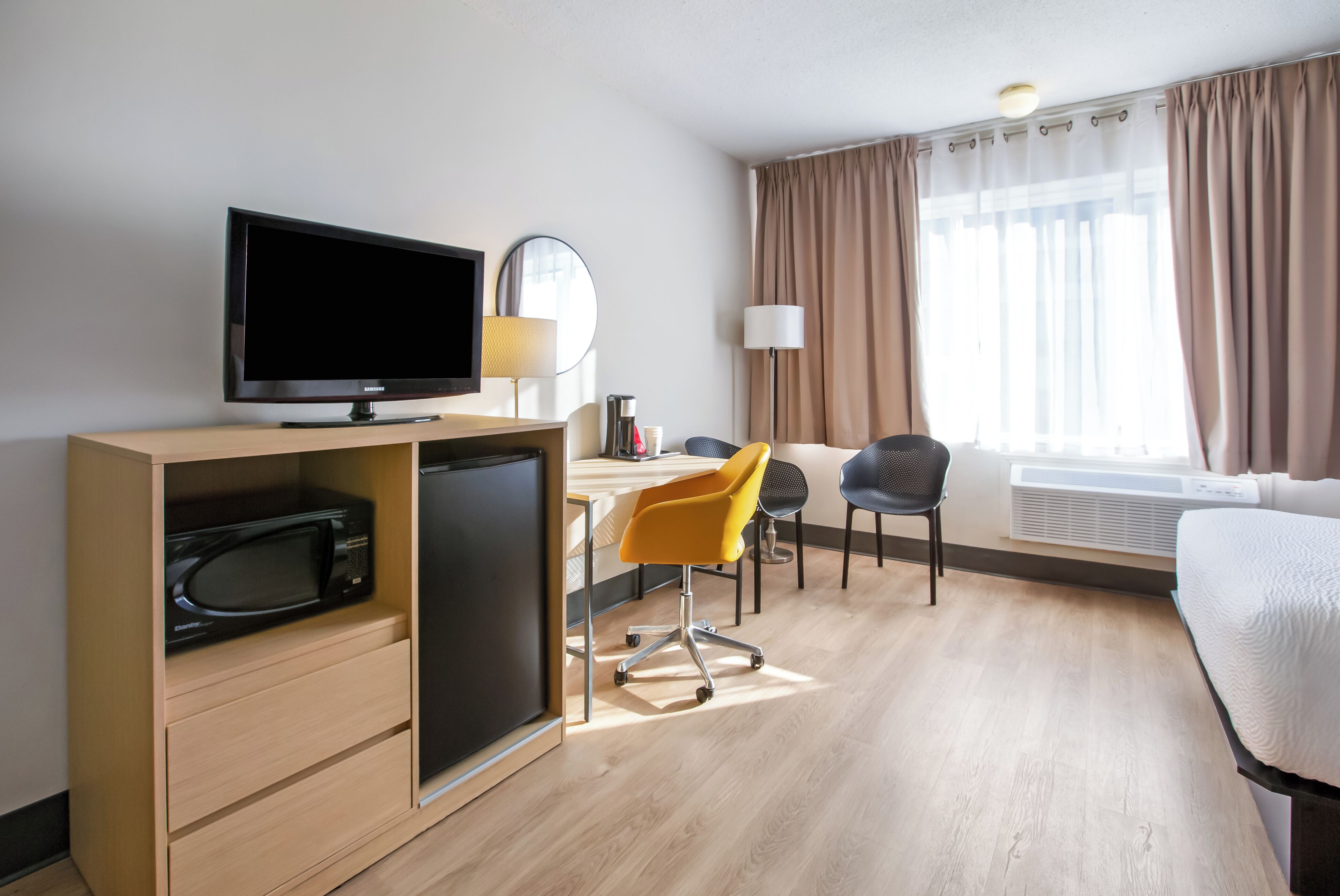 Quarto de hotel com televisão de tela plana, escrivaninha com luminária, cadeira, cama e janela com cortinas.
