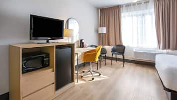 Quarto de hotel com televisão de tela plana, escrivaninha com luminária, cadeira, cama e janela com cortinas.