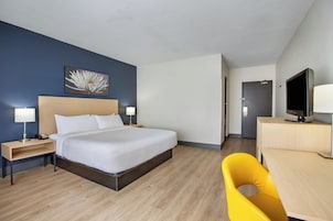 Quarto de hotel com cama, abajures, escrivaninha com cadeira, televisão e porta.