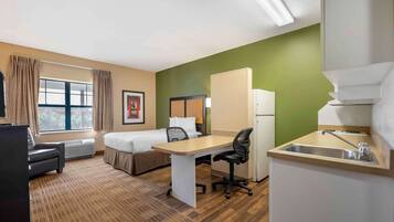Quarto de hotel compacto com cama, escrivaninha, cadeira, cozinha compacta e uma grande janela com cortinas.