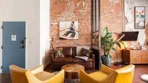 Uma sala de estar com parede de tijolos, sofá de couro marrom, cadeiras amarelas, mesa de centro e um vaso de planta, além de um quadro.