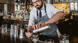 Um bartender prepara uma bebida num bar, com estantes cheias de garrafas ao fundo.