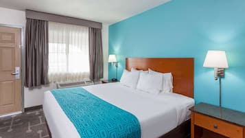 Quarto de hotel com cama, duas luminárias de cabeceira, mesa de apoio, janela com cortinas e porta.