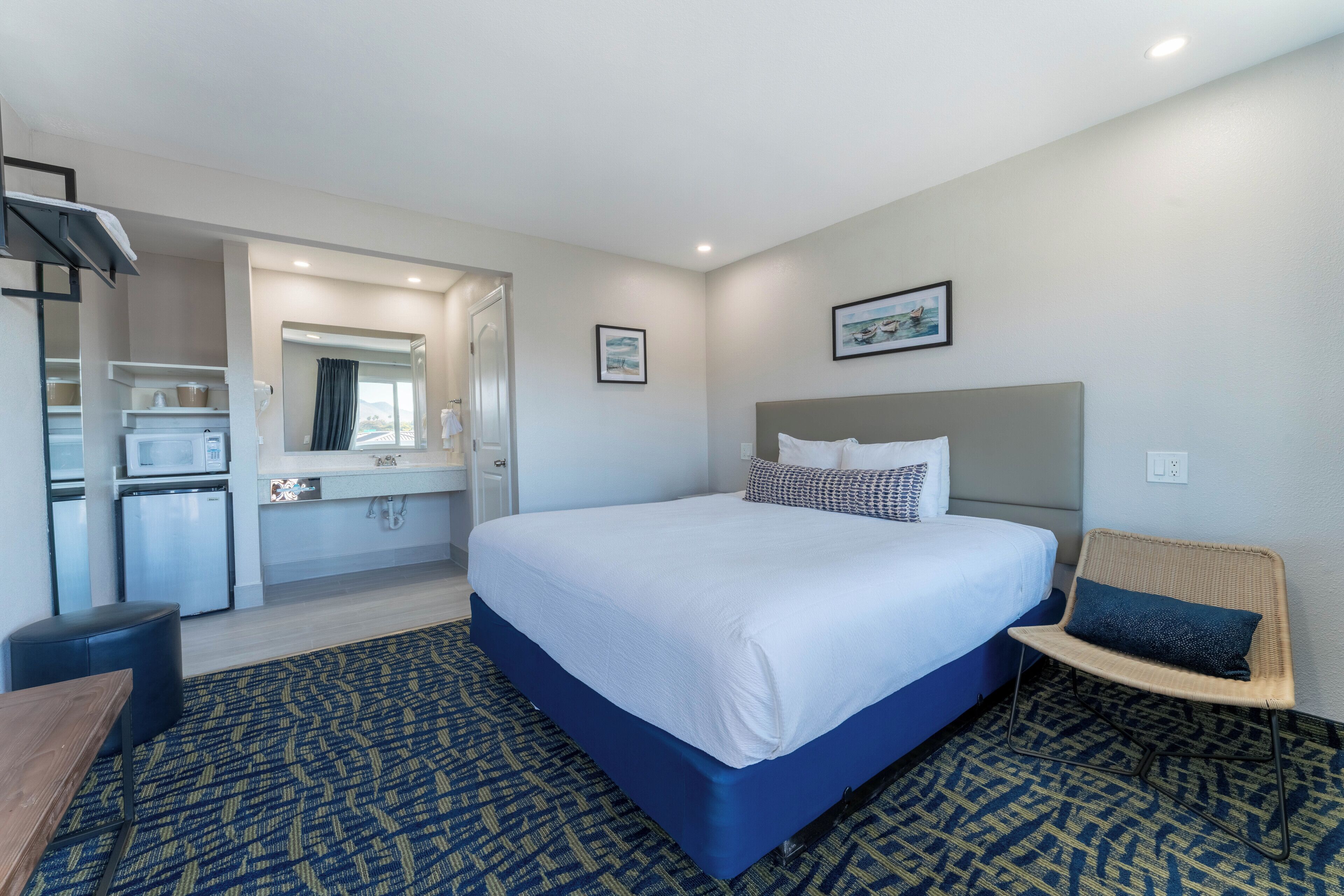 Quarto de hotel com cama, cadeira, escrivaninha, espelho e cozinha compacta.