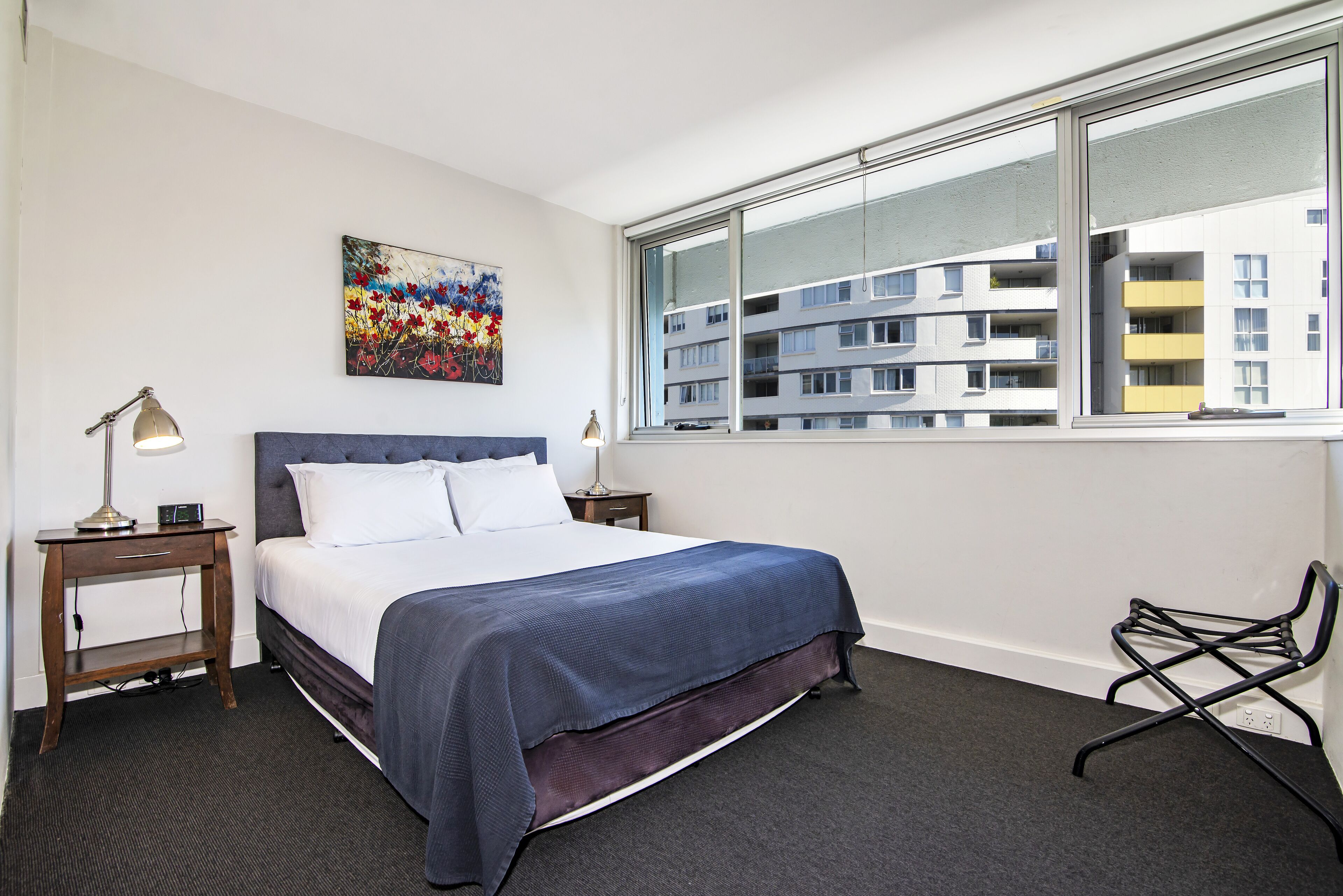 Quarto de hotel com uma cama grande, uma mesinha de cabeceira, uma luminária, um quadro e vista para um edifício pela janela.