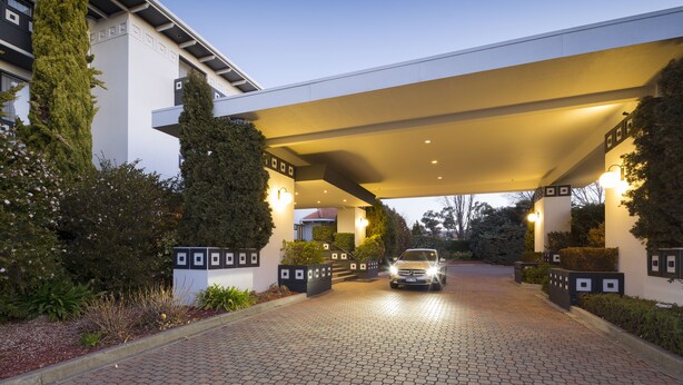 Entrada de hotel com calçada coberta, um carro estacionado em frente e um jardim bem cuidado.
