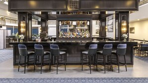 Uma área de bar com balcão de madeira escura, várias garrafas de bebidas e quatro banquetas.