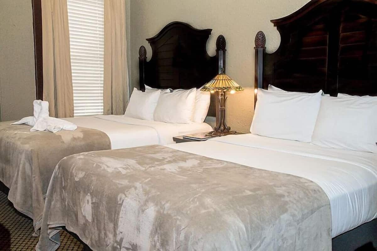 Quarto de hotel com duas camas, uma mesa de cabeceira com abajur e uma janela com persianas.