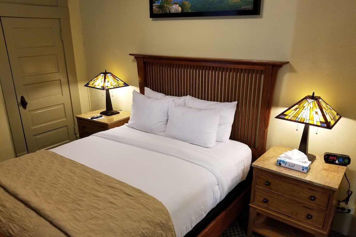 Quarto de hotel com uma cama, duas luminárias de cabeceira, um criado-mudo, um relógio digital e um quadro na parede.
