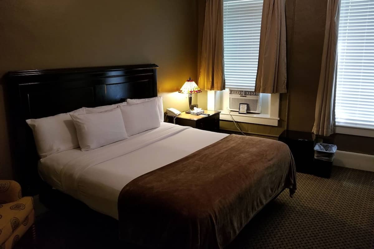 Quarto de hotel com cama, mesa de cabeceira, luminária, aparelho de ar condicionado e janela com cortinas.