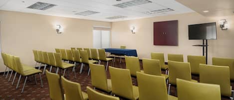 Uma sala de conferências com fileiras de cadeiras amarelas, uma mesa com toalha azul, uma televisão em um suporte e uma porta com janela de vidro deslizante.