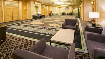 Um hall de hotel com piso carpeteado, área de estar com cadeiras, balcão de recepção e uma tela de televisão.