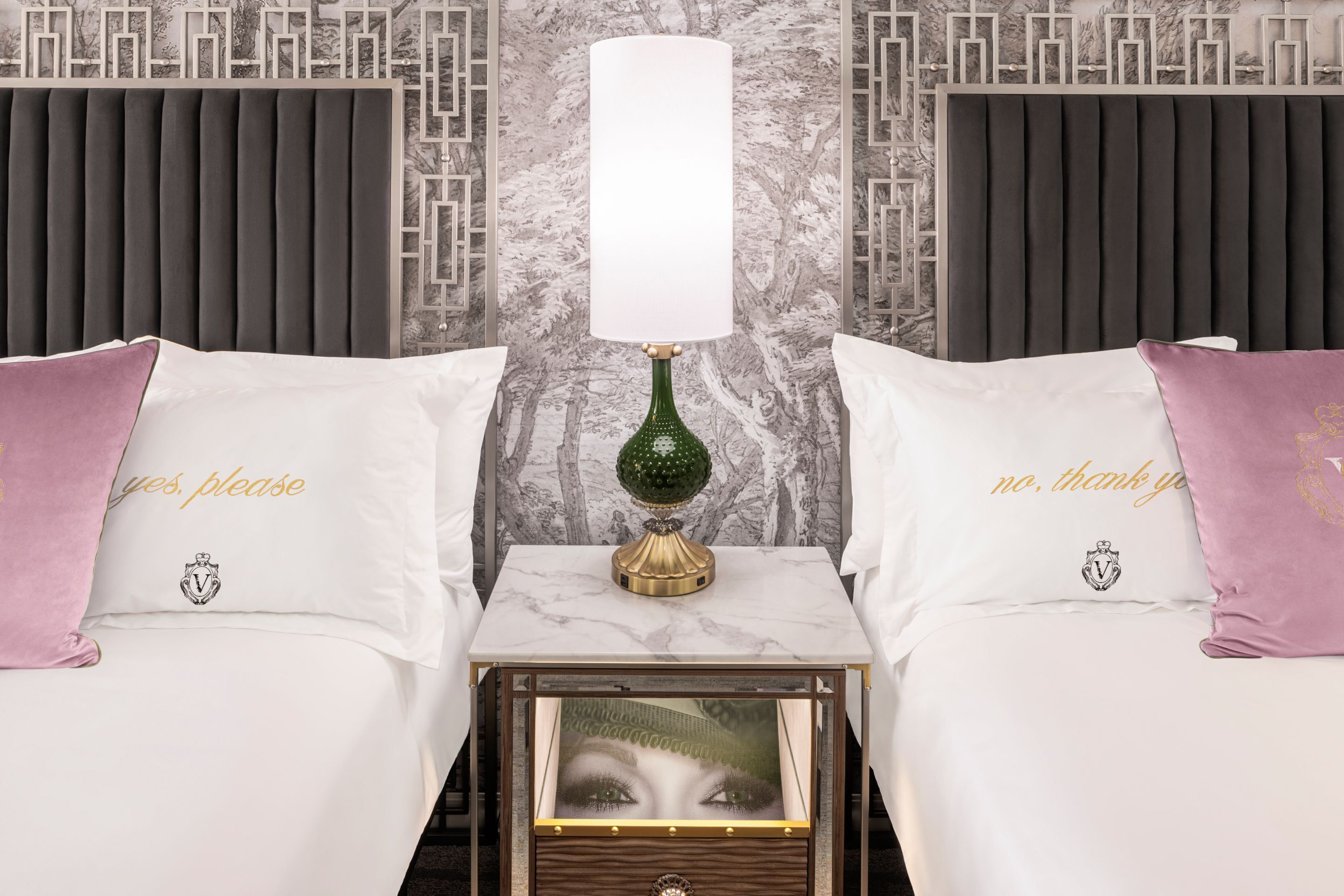 Quarto de hotel com duas camas, uma mesa de cabeceira com abajur e um painel decorativo.