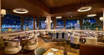 Restaurante com vista para o horizonte urbano noturno, com teto de madeira, mesas redondas e iluminação moderna.