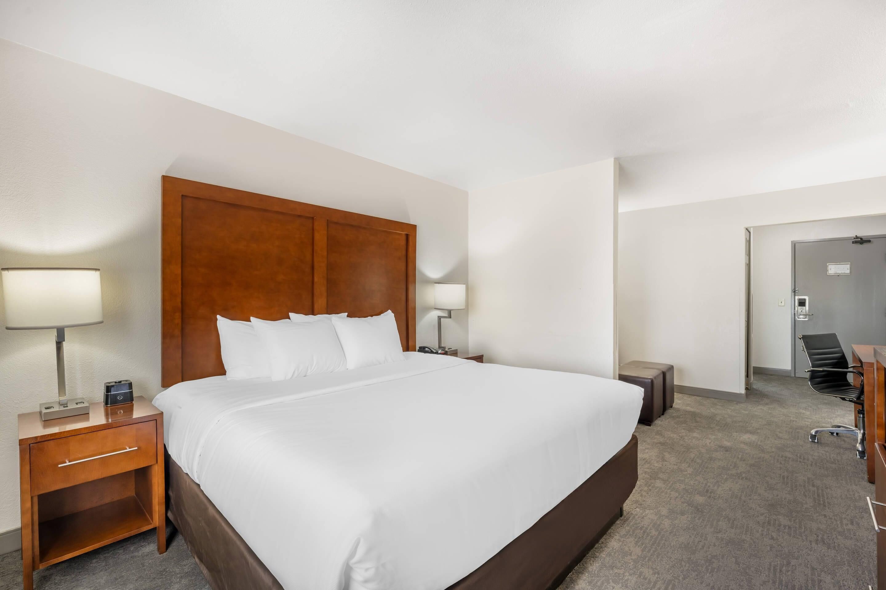Quarto de hotel com uma cama grande, duas mesas de cabeceira com luminárias, uma escrivaninha com cadeira e uma porta que dá para outro cômodo.