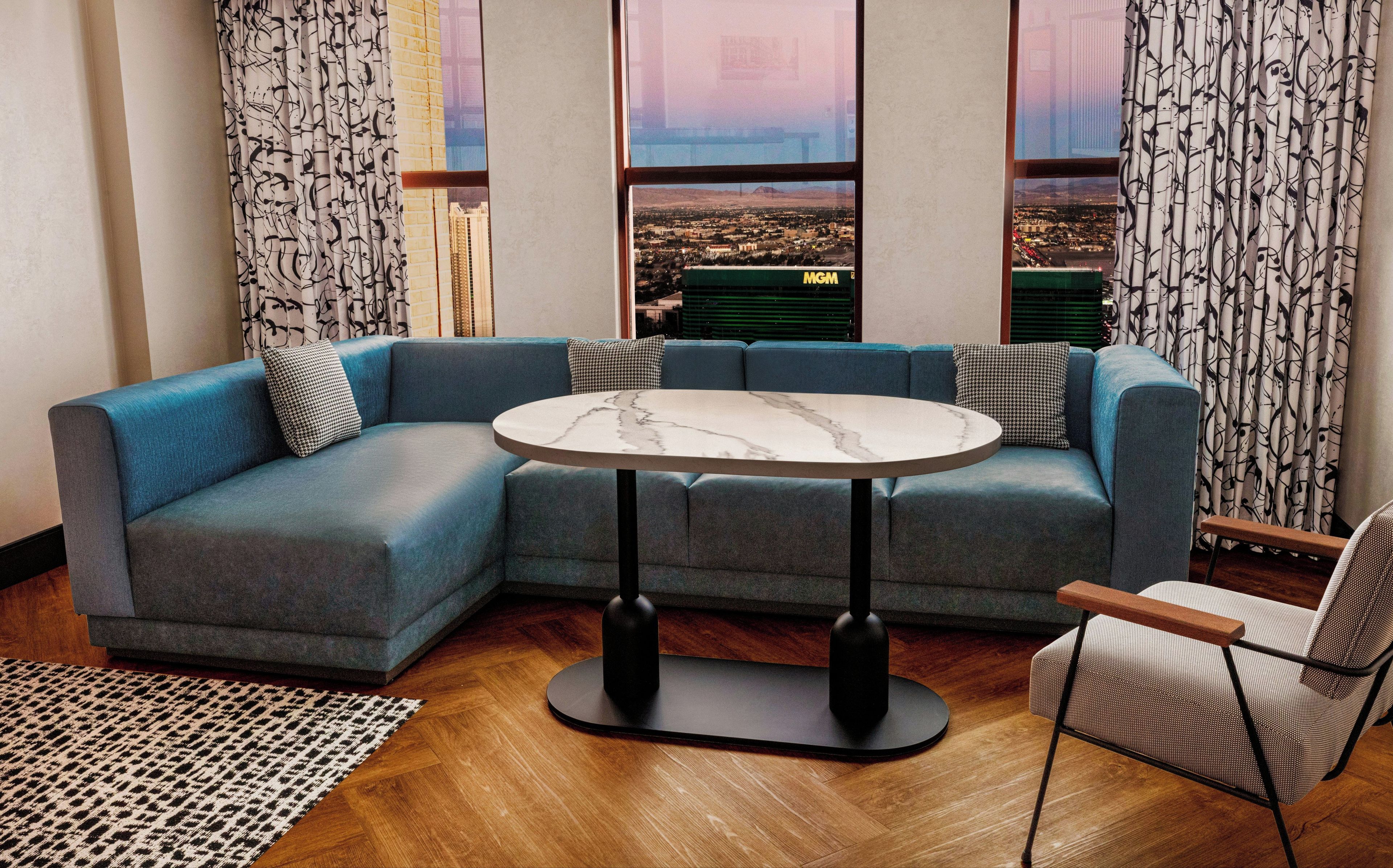 Uma sala de estar moderna com um sofá seccional, uma mesa redonda e vista para o horizonte urbano por amplas janelas.