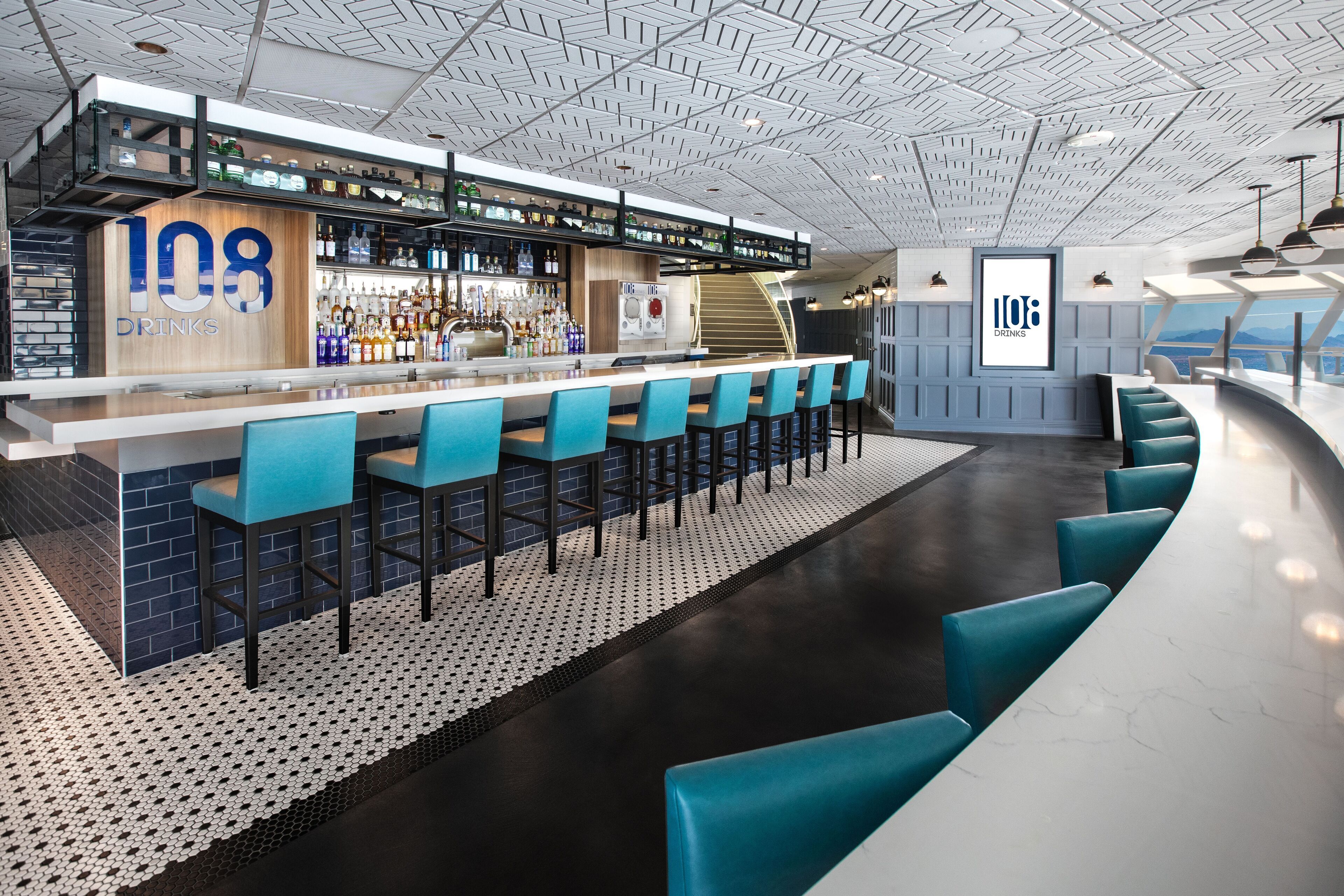 Uma área de bar moderna com um balcão exibindo '108 DRINKS', vários bancos e uma escada que leva a um andar superior.
