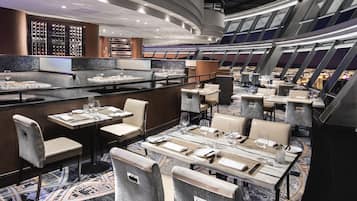 Restaurante moderno com pé direito alto e vista panorâmica da cidade. Mesas bem dispostas com copos de vinho e talheres.