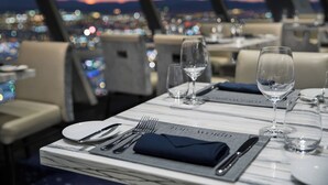 Um restaurante com mesa posta para refeição, vista para a cidade ao fundo e um pôr do sol sobre as montanhas.